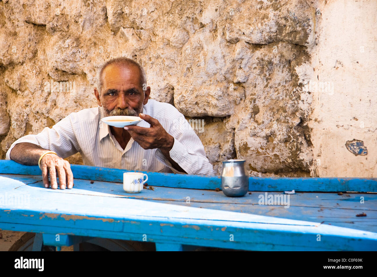 Old Indian man drinking coffee on a blue wooden table in the street ...