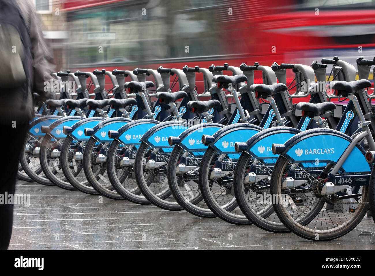 A rack of Boris Bikes, a blurred red London bus passing in the background and a person walking ...