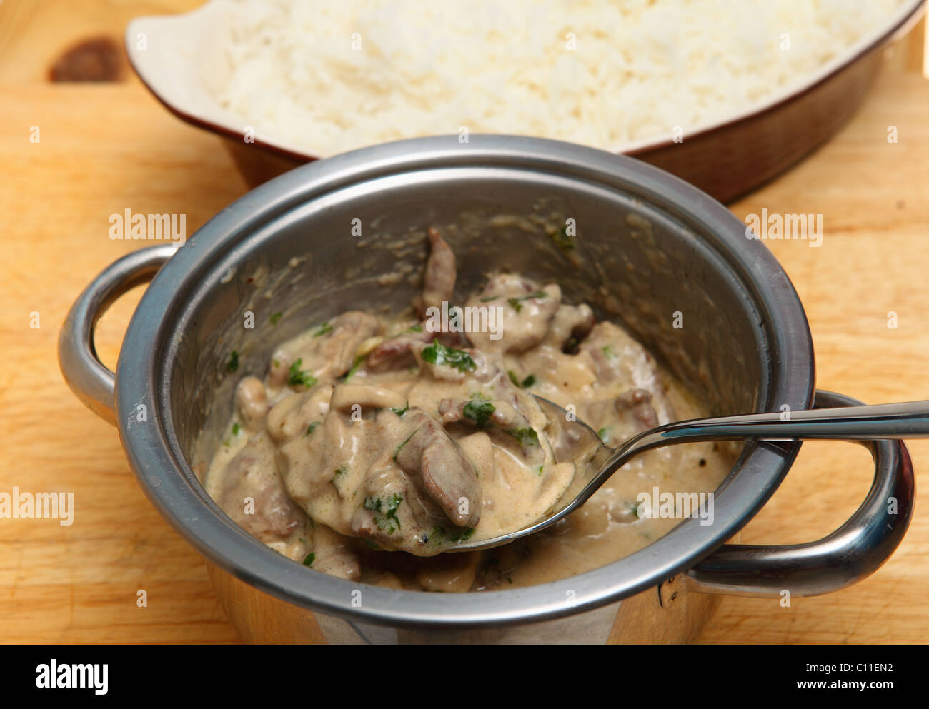 A cooking pot full of beef stroganoff with a serving spoon and a bowl ...