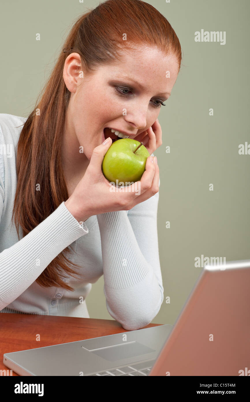 Long red hair woman biting apple at office watching laptop screen Stock ...