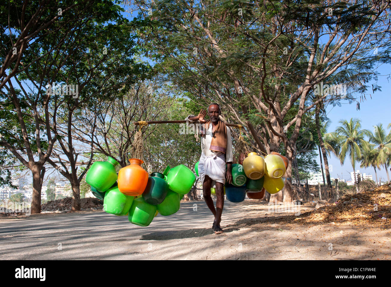 Indian man carrying plastic water pots along a tree lined road. Andhra ...