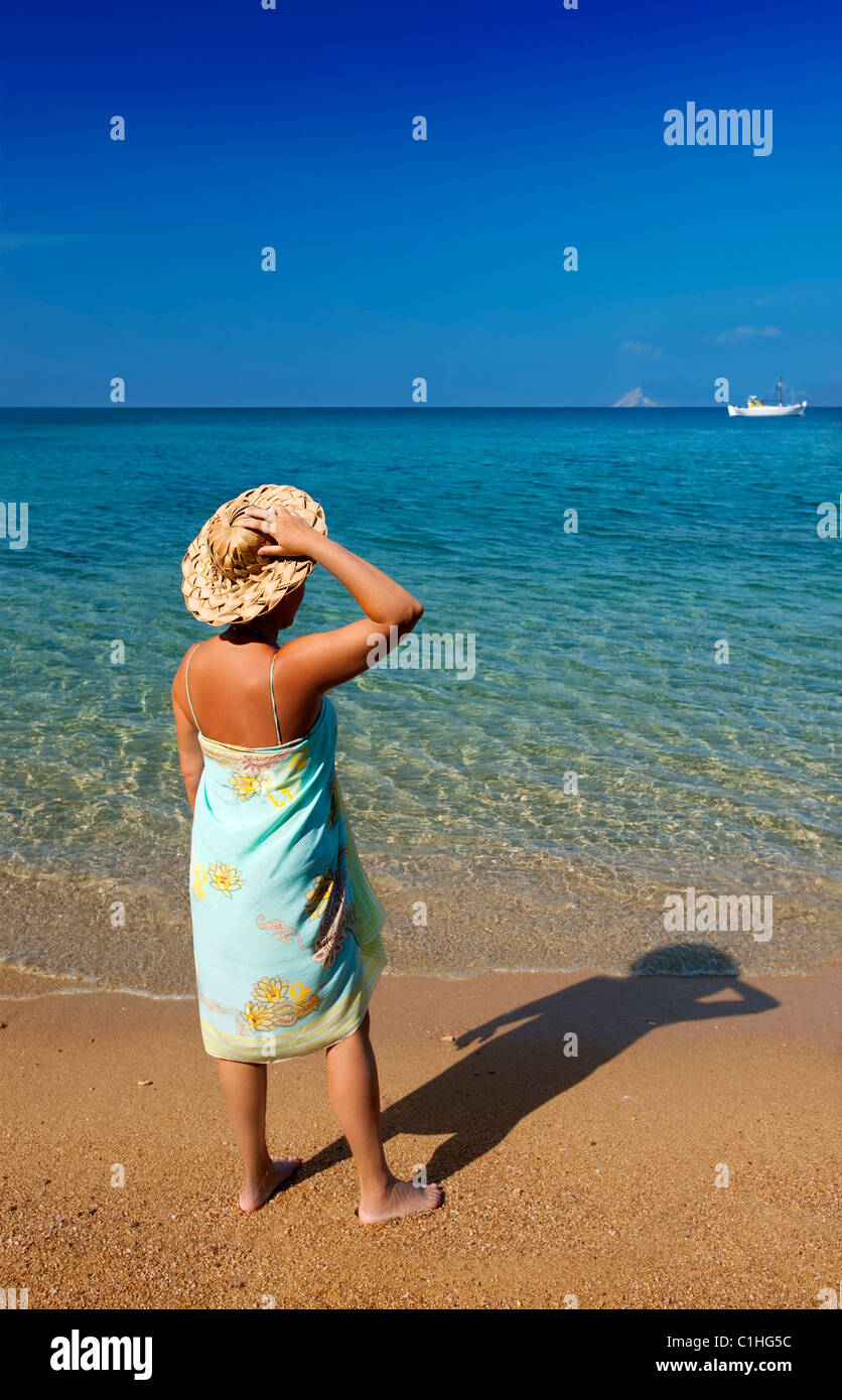 Young woman in a pareo and straw hat, standing on a Mediterranean sandy ...