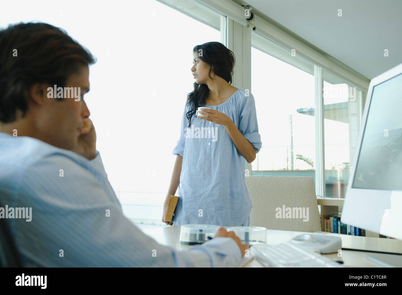 Woman standing with cup of coffee, looking out window, husband working ...