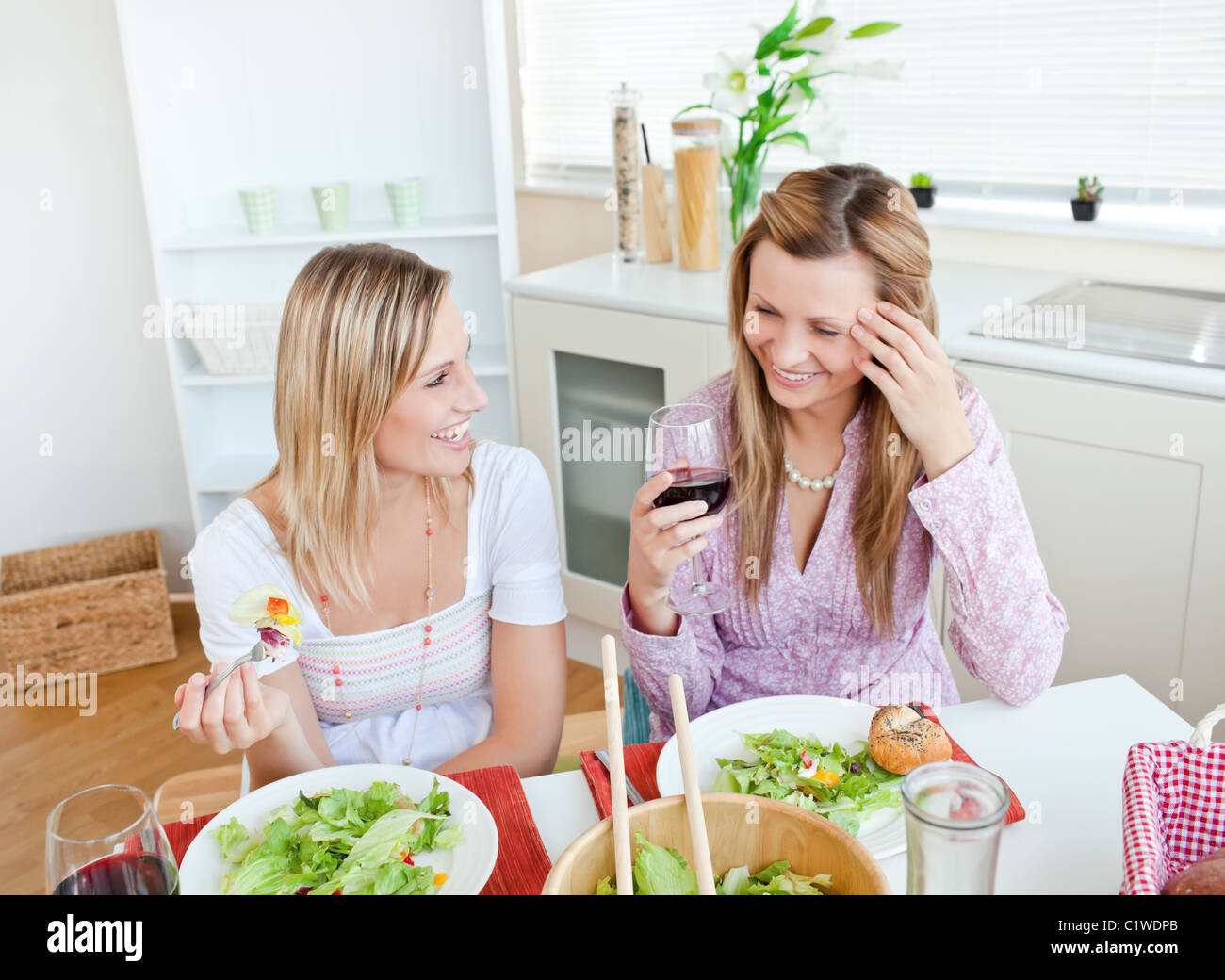 Two women in the kitchen chatting and eating salad with glasses of red ...