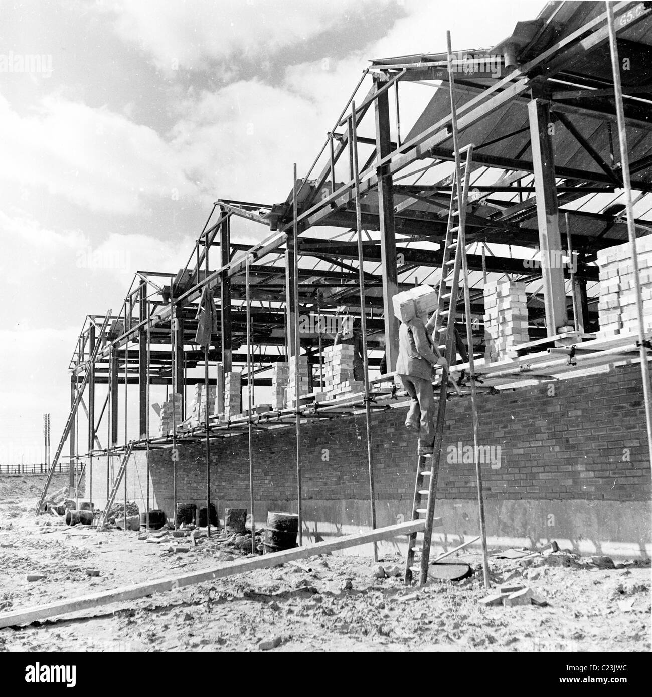 1950s, England. Bricklayer climbs a wooden ladder with bricks on his ...