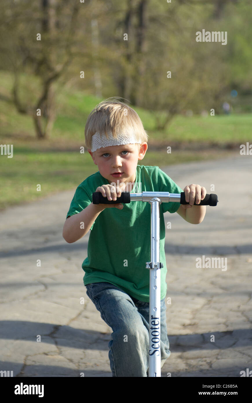 A young child blond boy on a scooter in park with injured head ...