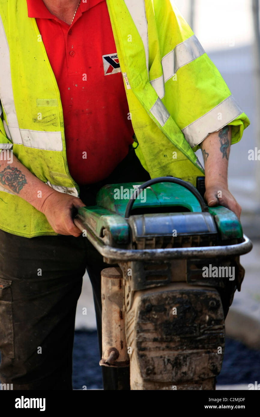 Road repair workman using a vibrating compactor to squash tarmac into a ...
