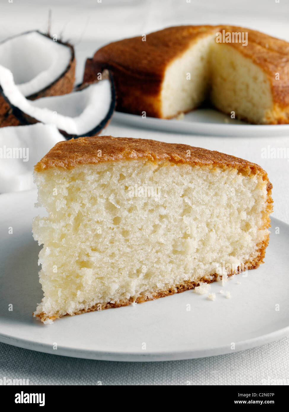 A slice of coconut cake with a whole cake and coconut pieces behind ...