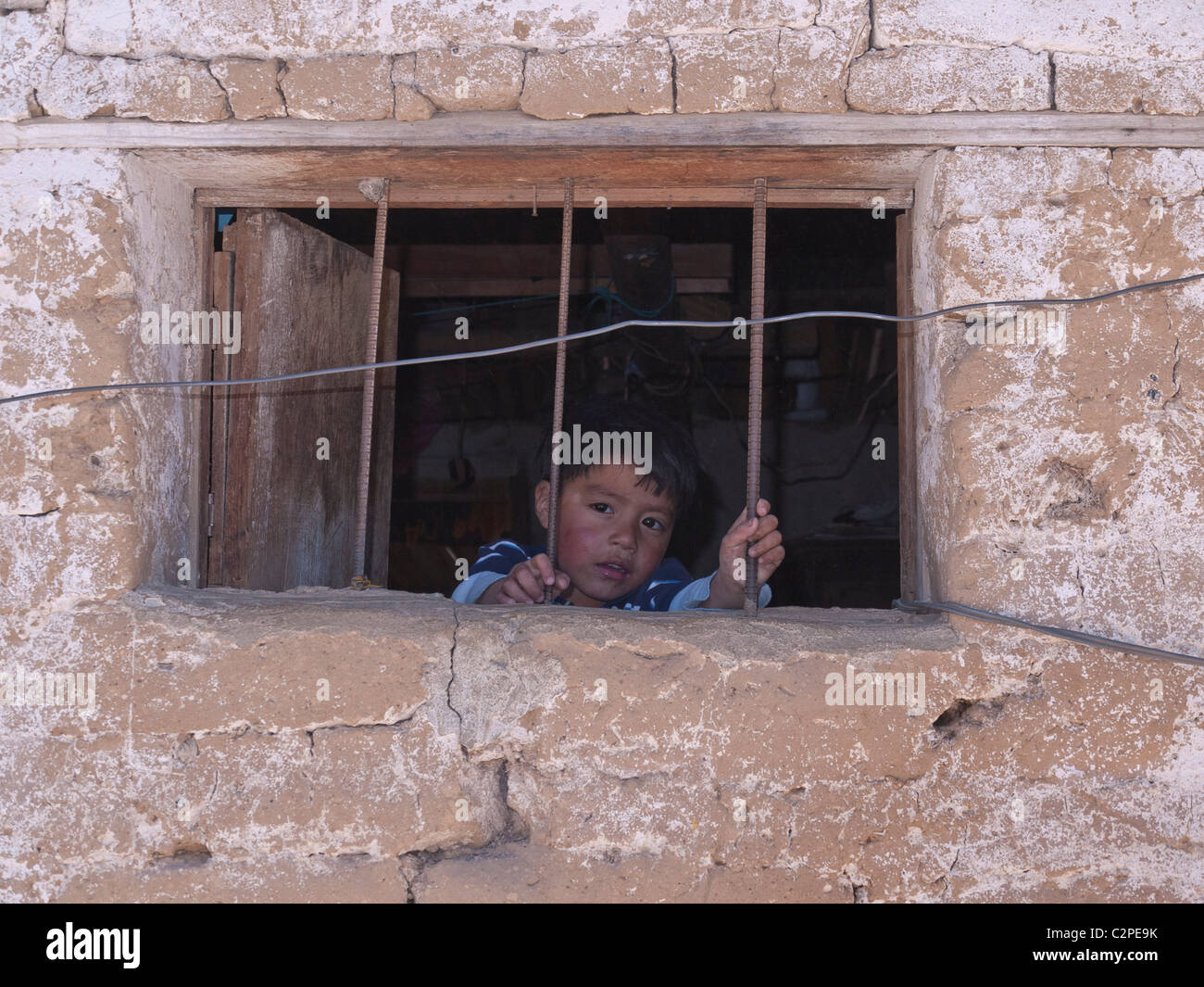 A very young boy looks out of the barred window of his adobe home and ...