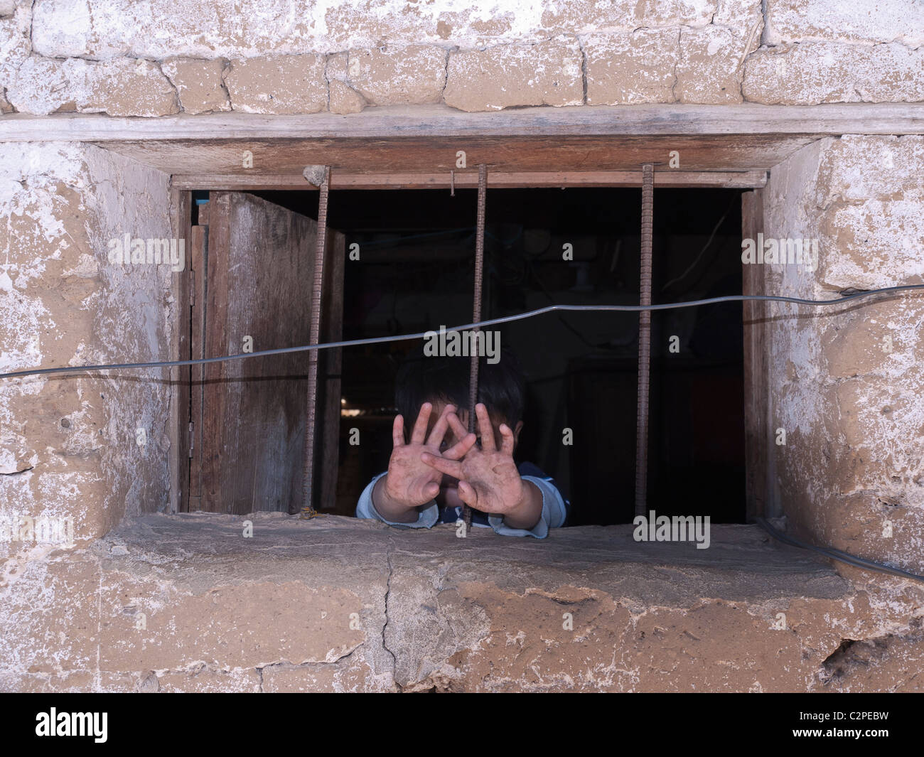 A very young boy looks out of the barred window of his adobe home and ...