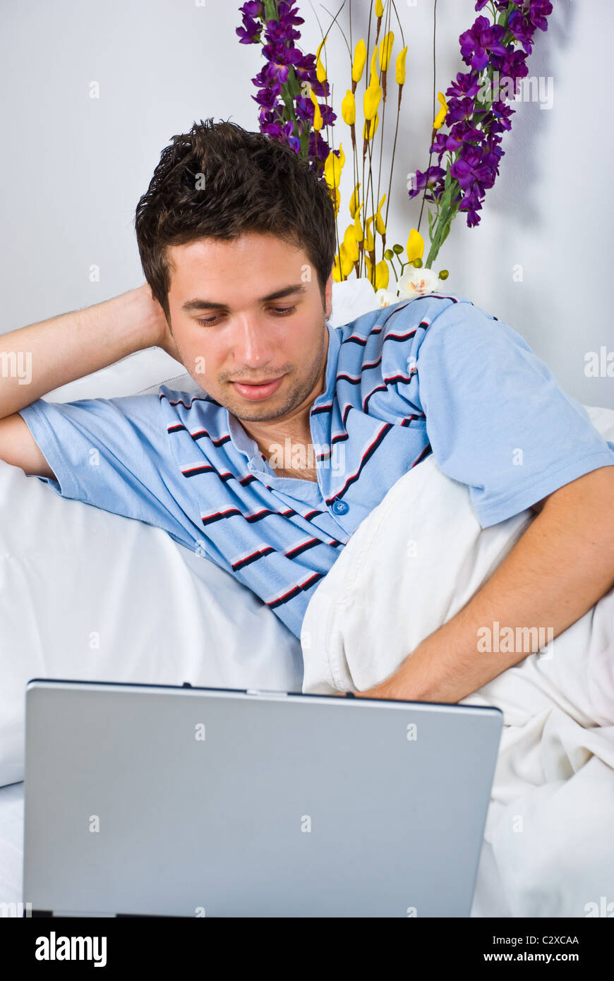 Young man sitting on bed in pajamas and browsing internet on laptop ...