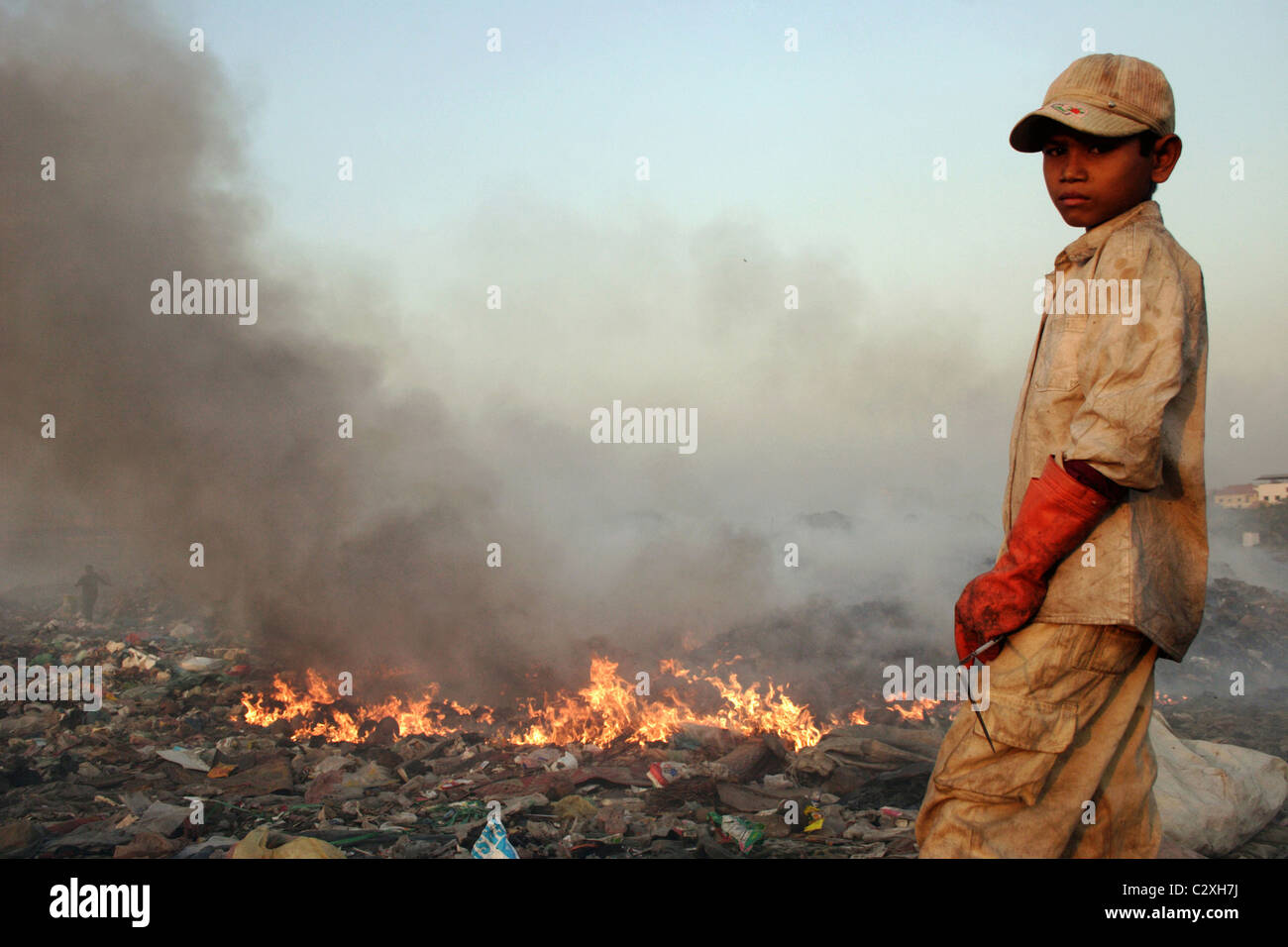 A young child laborer boy is standing near a pile of burning garbage in ...