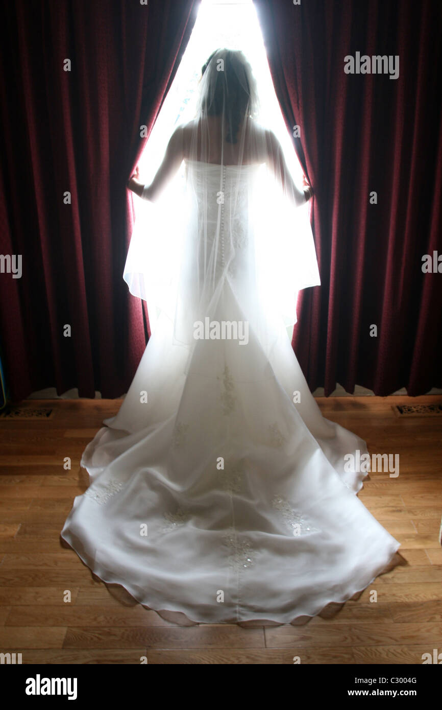 A vertical picture of a bride parting dark curtains to look out a ...