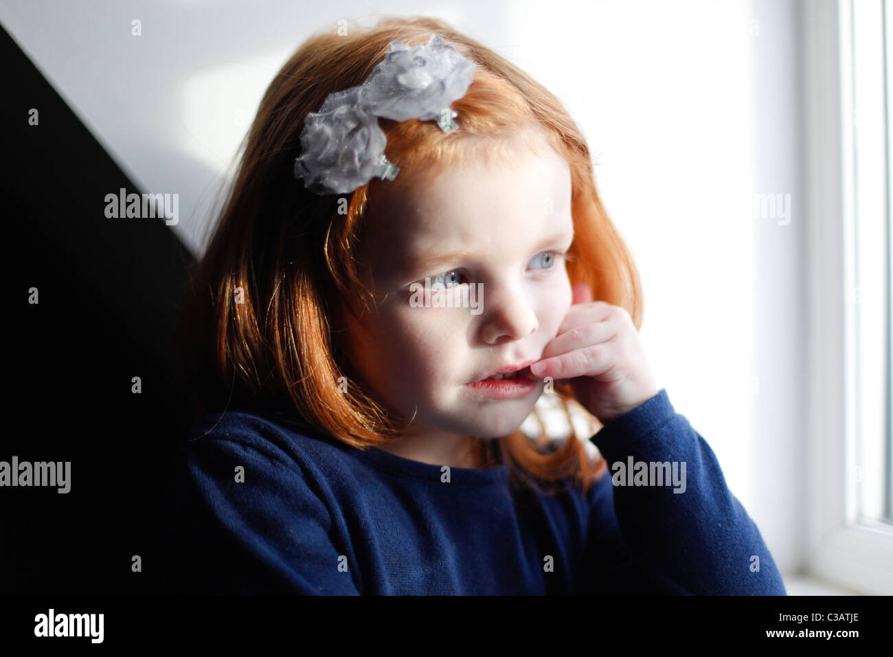 A four year old girl with red hair looking out of the window pondering ...