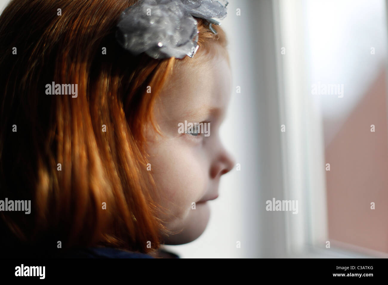 A four year old girl with red hair looking out of the window pondering ...