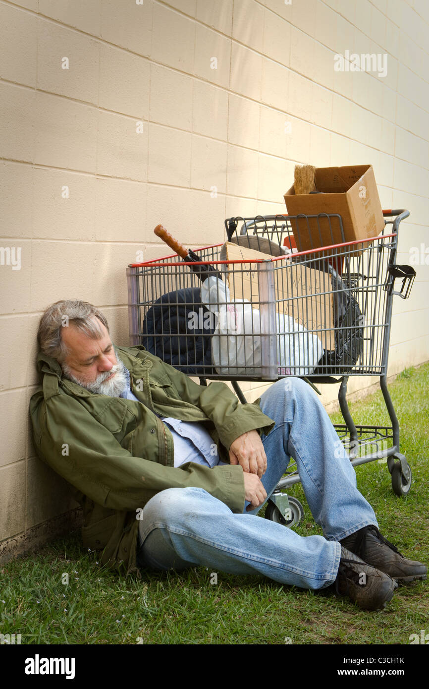 Homeless man leans against a wall sleeping with a grocery basket next ...
