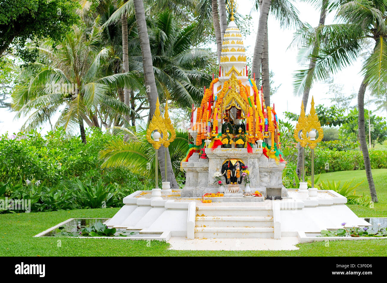An alabaster Thai altar sits colorfully decorated in a garden of green ...