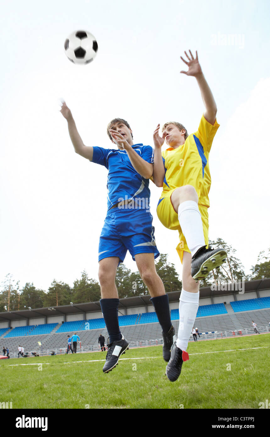 Two footballers jumping and looking at ball on grass-field during game ...