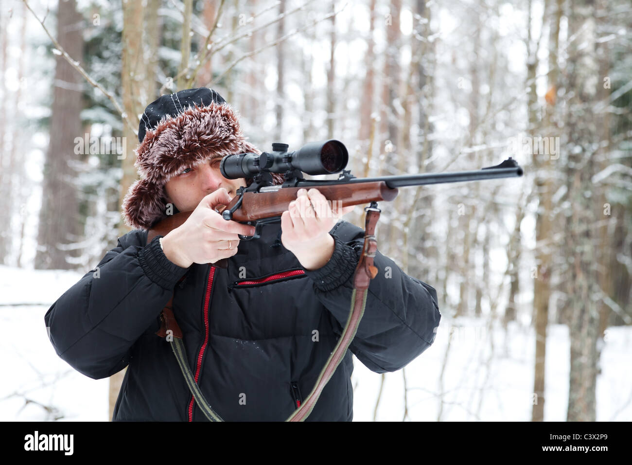 Hunter in a fur cap with ear flaps with sniper rifle in winter forest ...