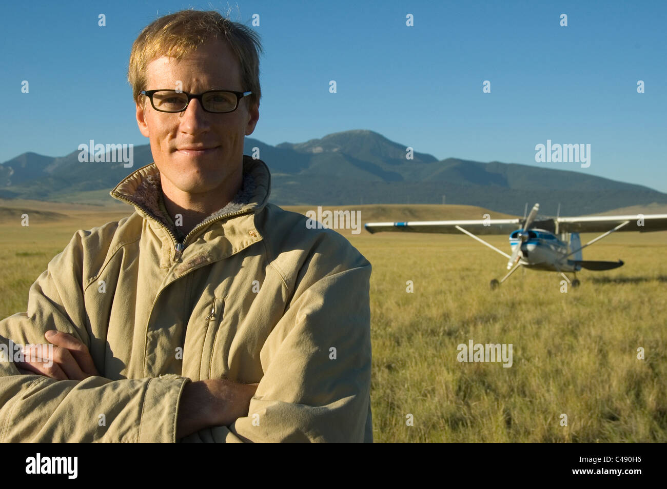 A man stands in a field next to his airplane with mountains in the ...
