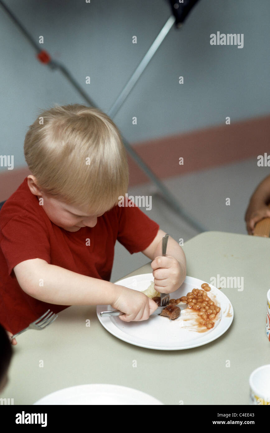 Nursery school boy eating school dinner of baked beans and sausage ...