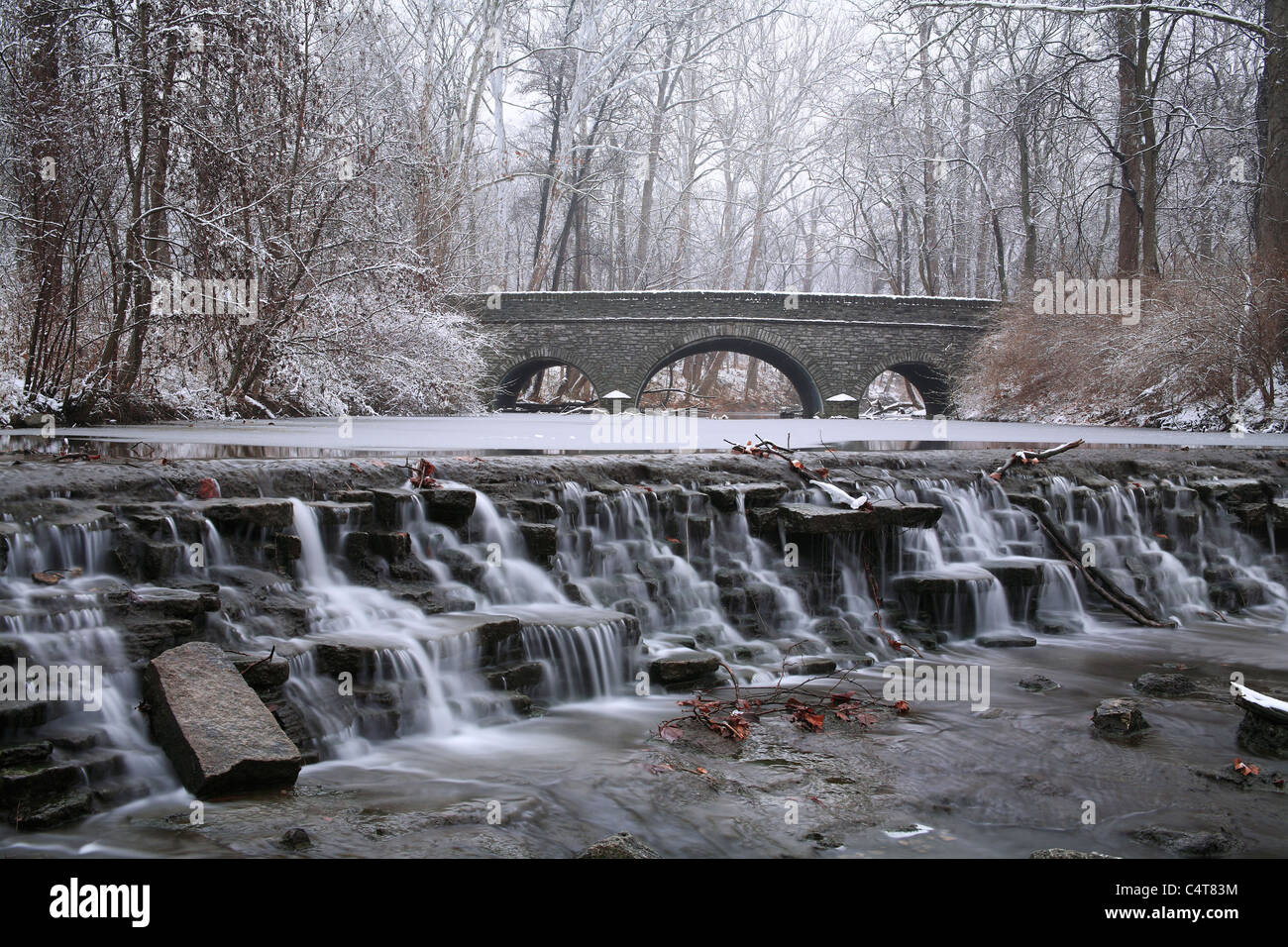 Snow Covered Trees Framing A Stone Bridge And Waterfall During Winter ...