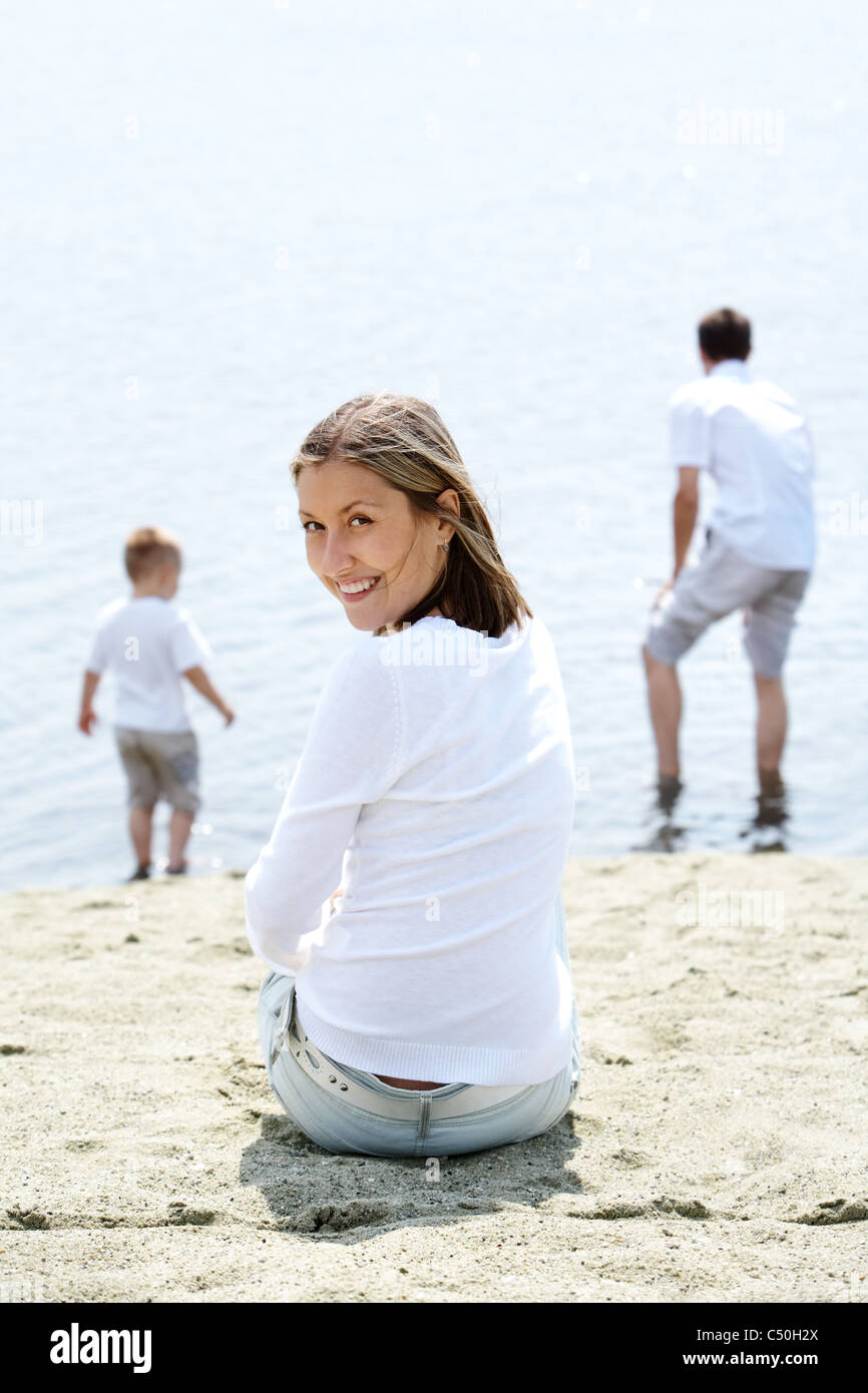 Image of smiling female sitting on sandy shore and looking at camera ...
