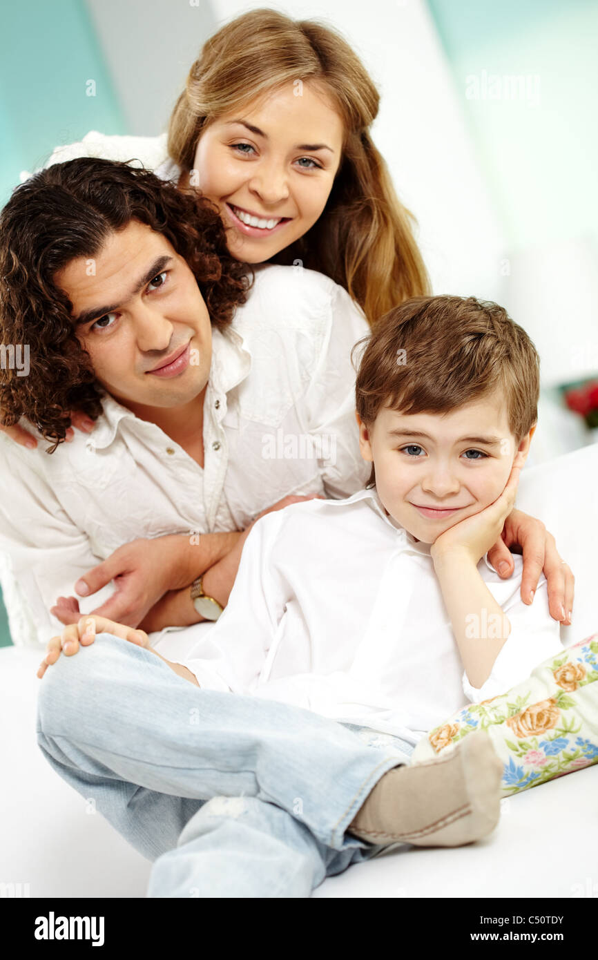 Cute lad sitting on sofa and looking at camera with his parents near by ...