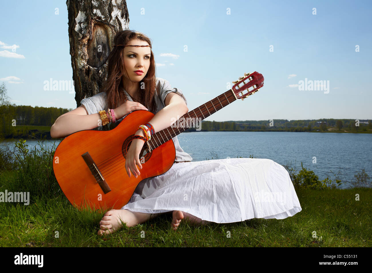 outdoor portrait of beautiful hippie girl sitting on green grass near birch with guitar. lake ...