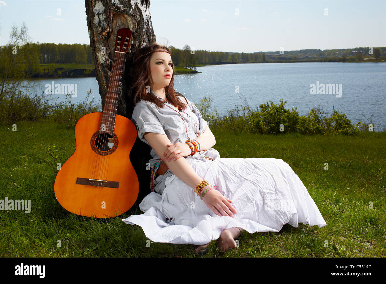 outdoor portrait of beautiful hippie girl sitting on green grass near birch with guitar. lake ...