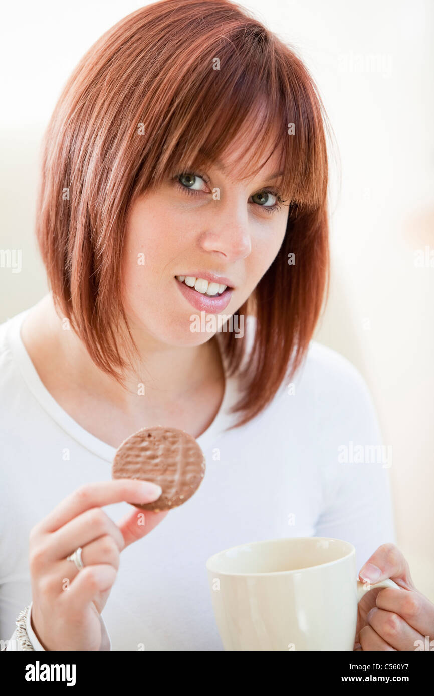 Attractive young woman eating a chocolate biscuit and drinking a mug of ...