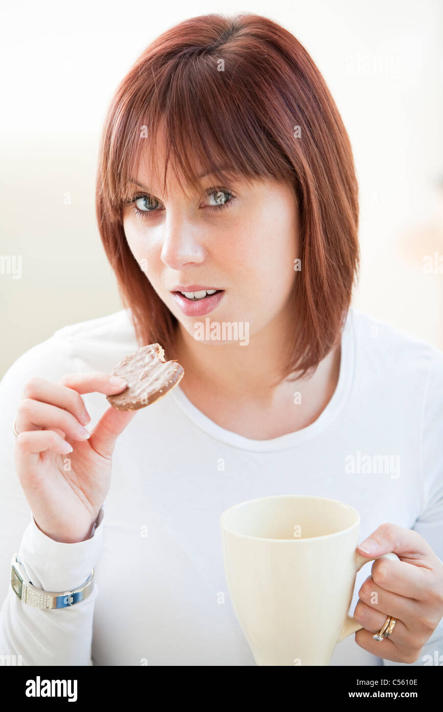 Attractive young woman eating a chocolate biscuit and drinking a mug of ...