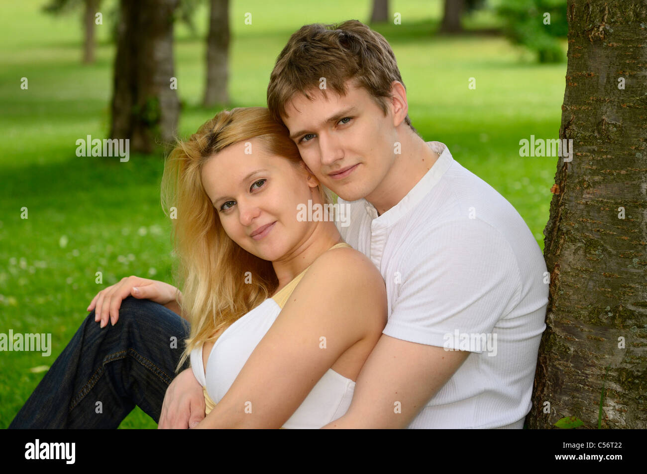 Cuddling white engaged couple sitting by a tree in a park Stock Photo ...
