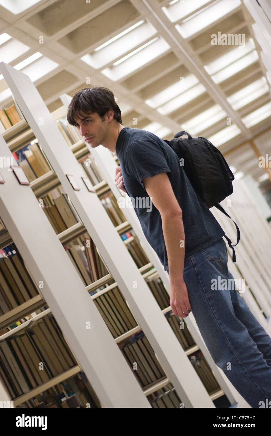 A young man reads a book at the library while standing in the aisles of ...