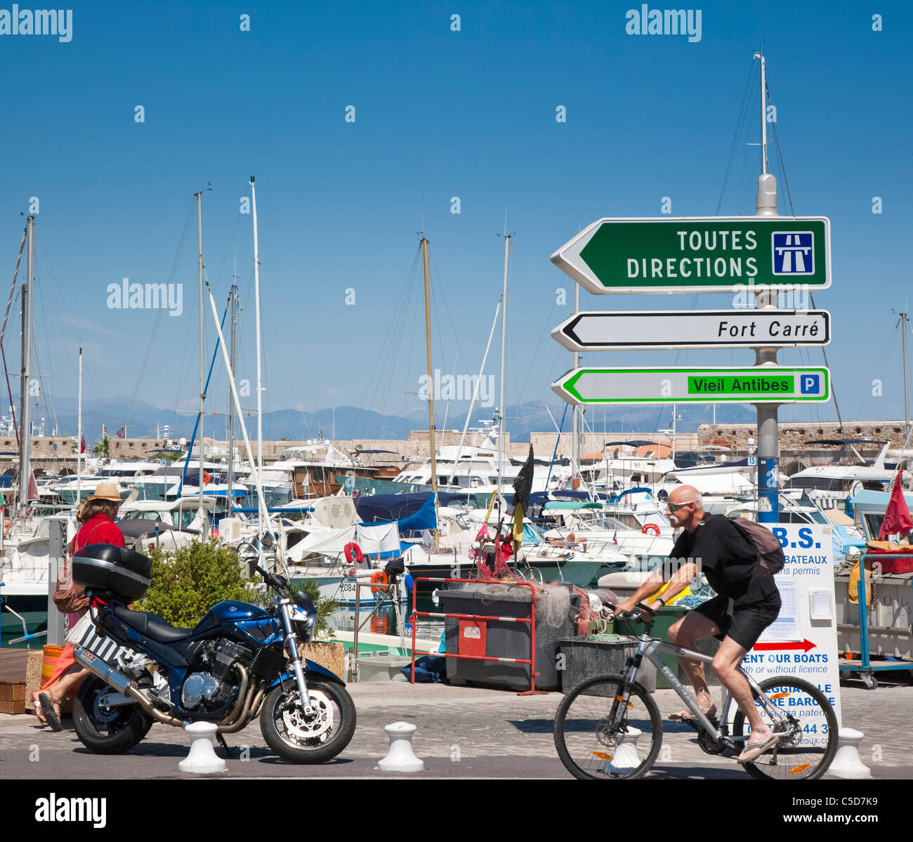 Cyclist and motorbike with direction sign and boats behind, Antibes ...