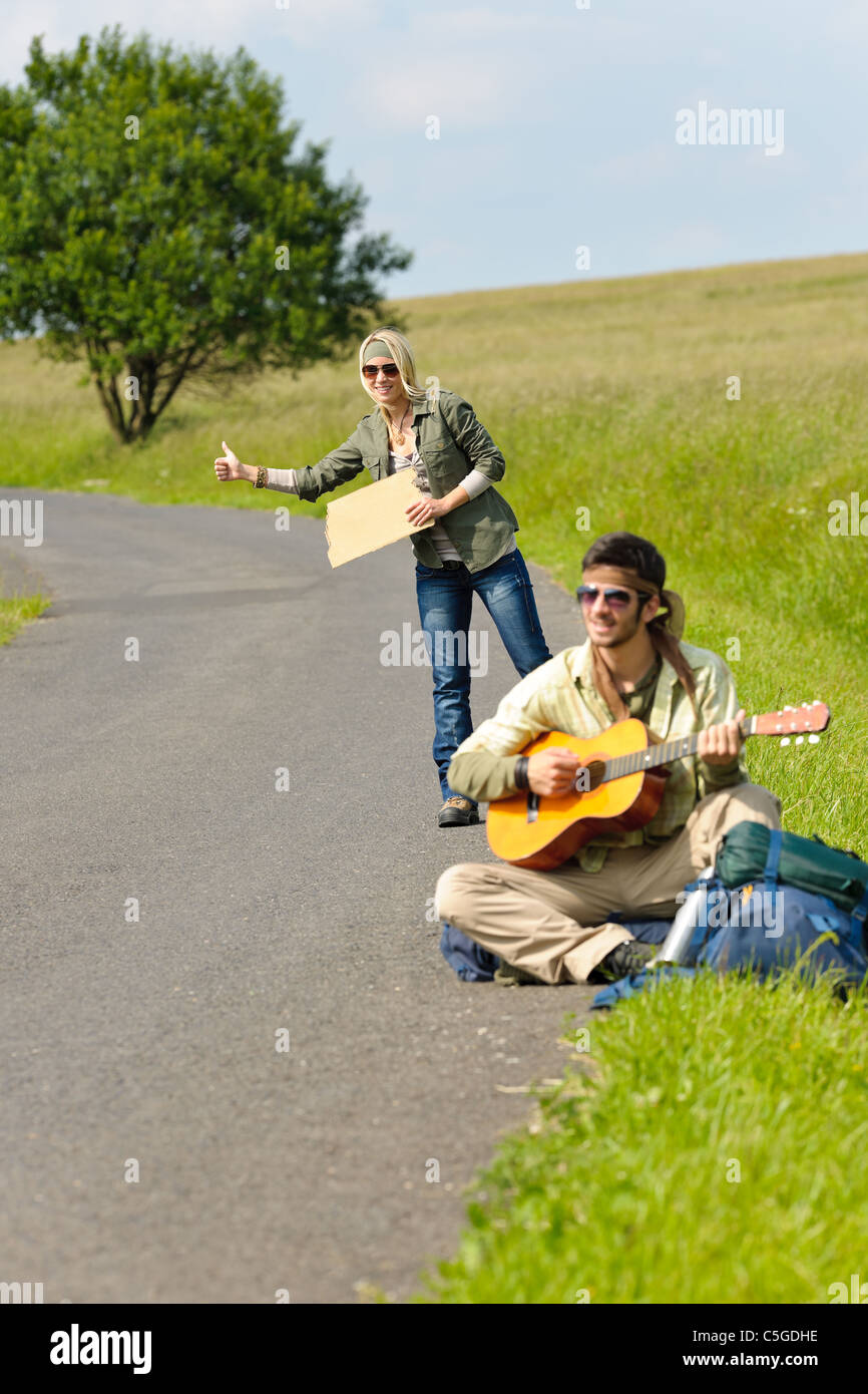 Hitch-hike young couple backpack tramping on asphalt road play guitar ...