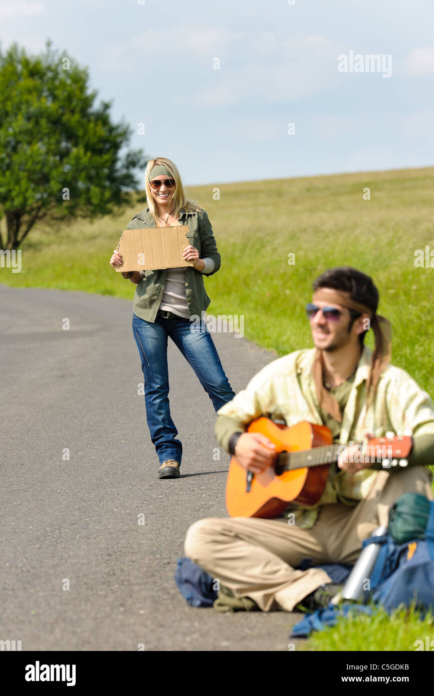 Hitch-hike young couple backpack tramping on asphalt road play guitar ...