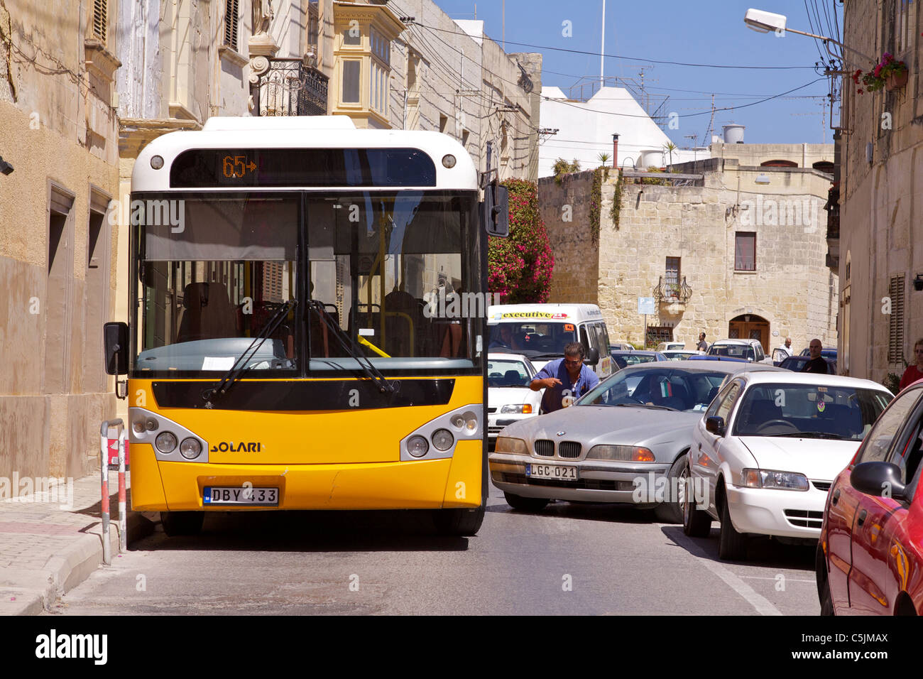 A yellow Maltese bus attempts to squeeze past a broken down car in a ...
