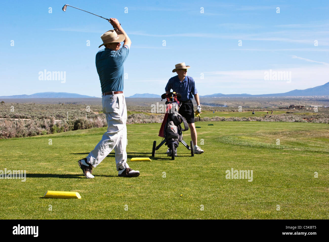Two golfers on golf course, one has hit the ball and the other watches ...