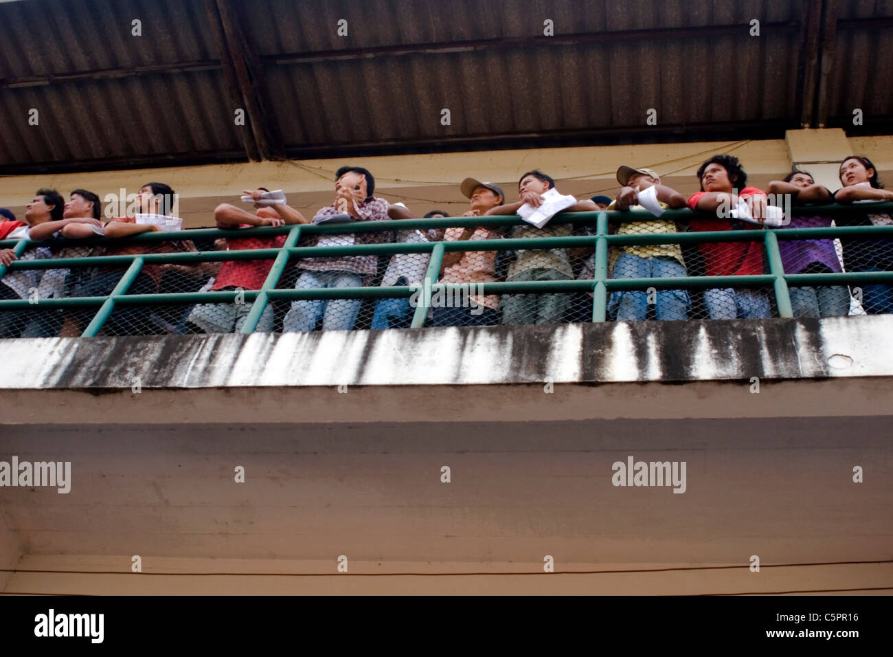 Horse racing fans are leaning on a balcony rail watching a horse race ...