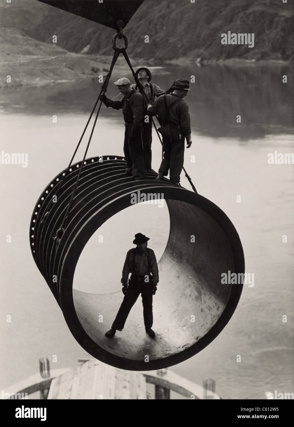 Four workers riding on a large casing section of pipe suspended from ...