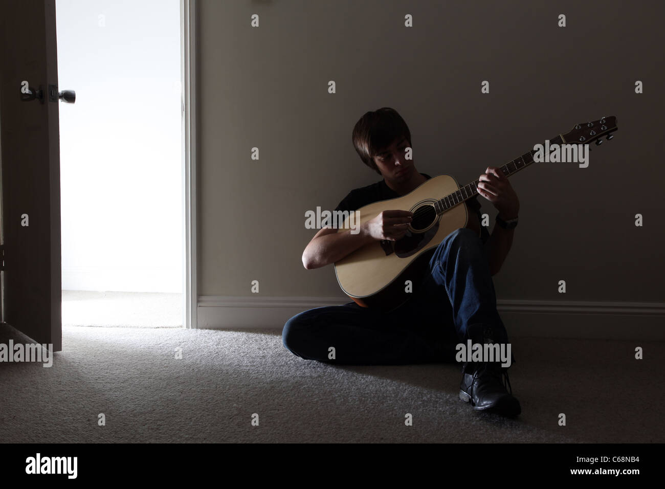 Young man sitting alone playing an acoustic guitar in a dark room with ...