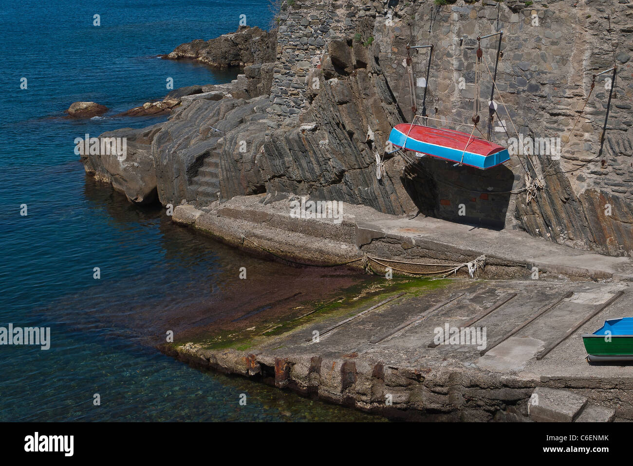 A small boat is hanging by ropes from a rock cliff by water's edge in a ...