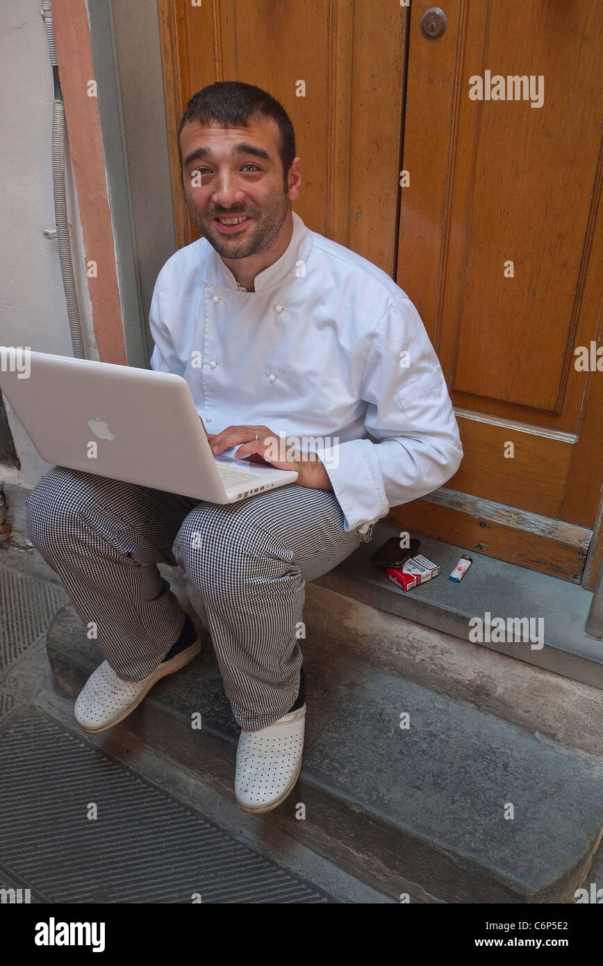 A young Italian male chef works on his laptop on the front step outside ...