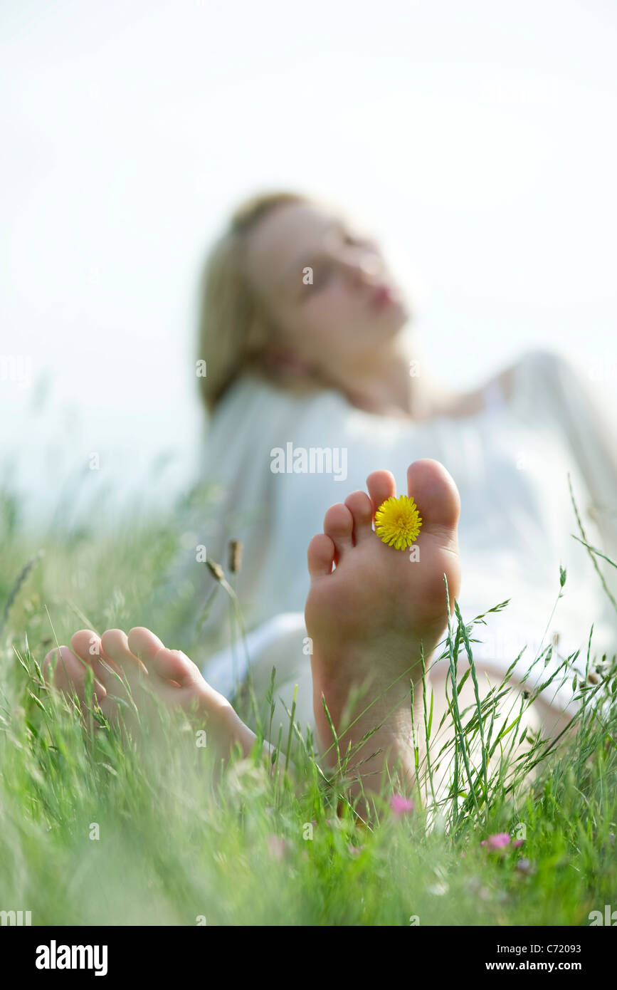 Barefoot young woman sitting in grass, holding dandelion flower between ...
