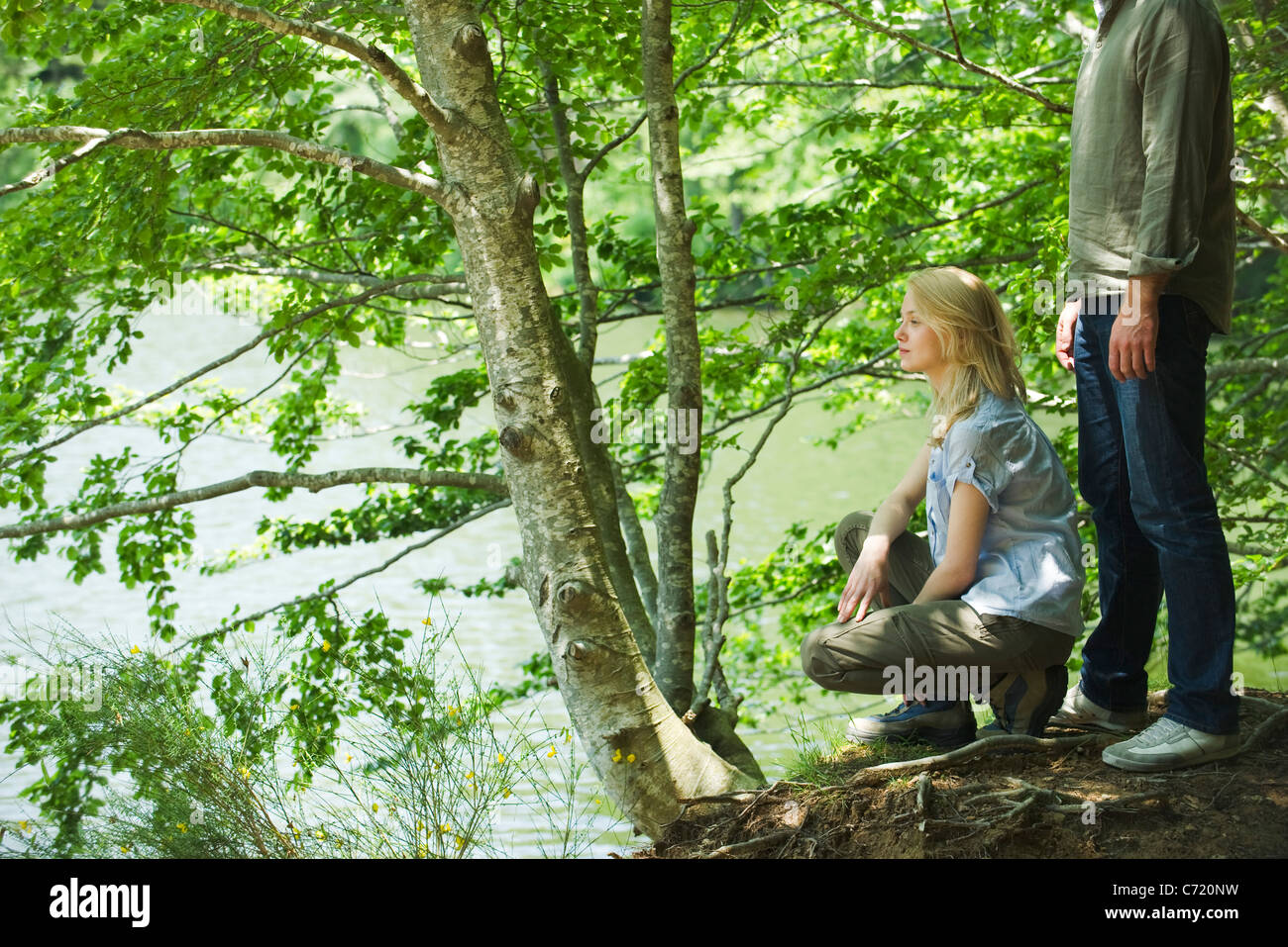 Young woman crouching beside man, looking at lake Stock Photo - Alamy