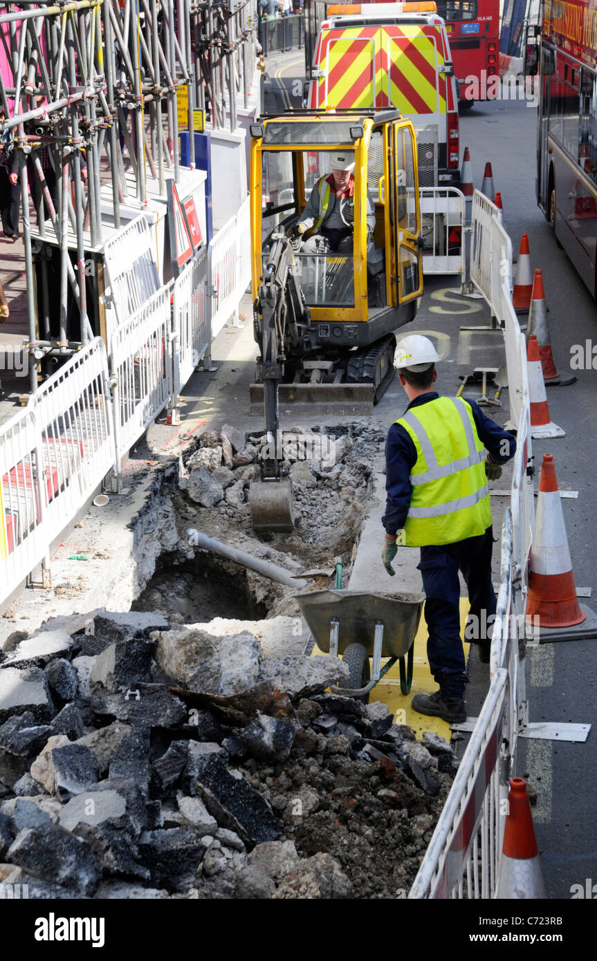 Looking down on mini digger excavator & worker in narrow space for road ...
