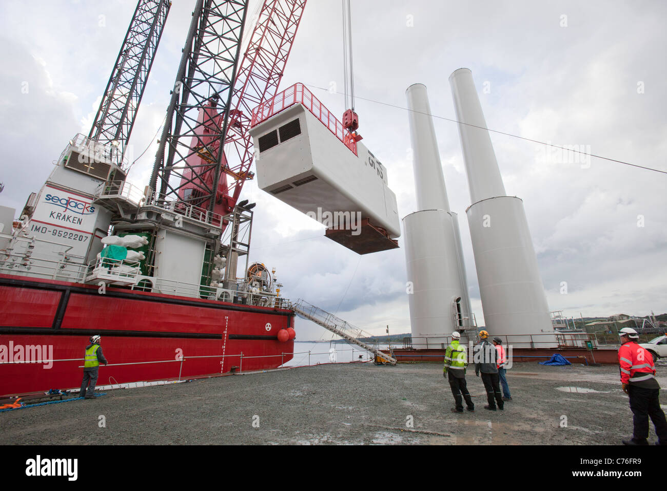 Workers lift a nacelle for a wind turbine onto a jack up barge, for the ...