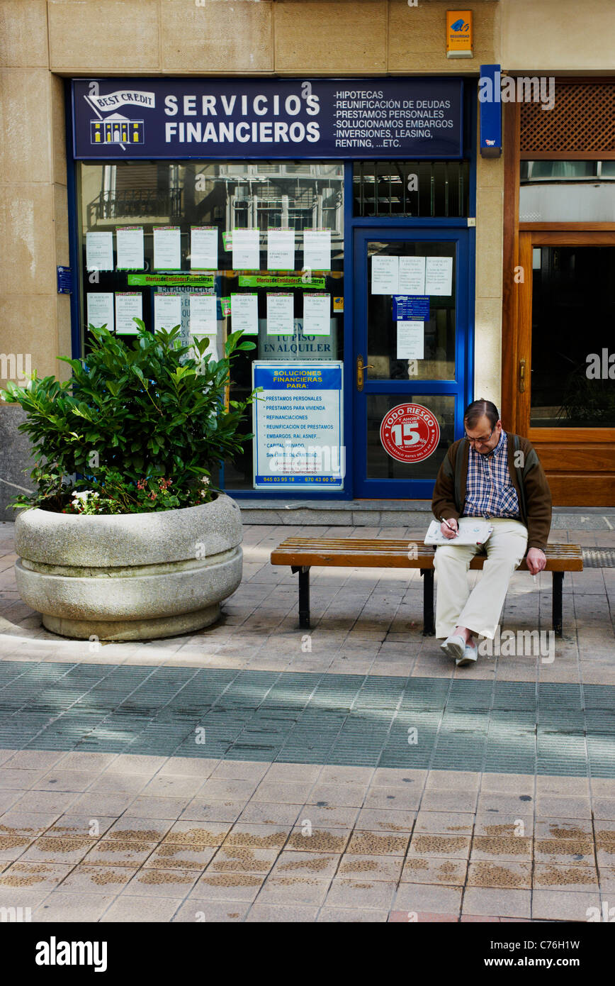 A man sits on a bench smoking, whilst he writes on some paper, sitting ...