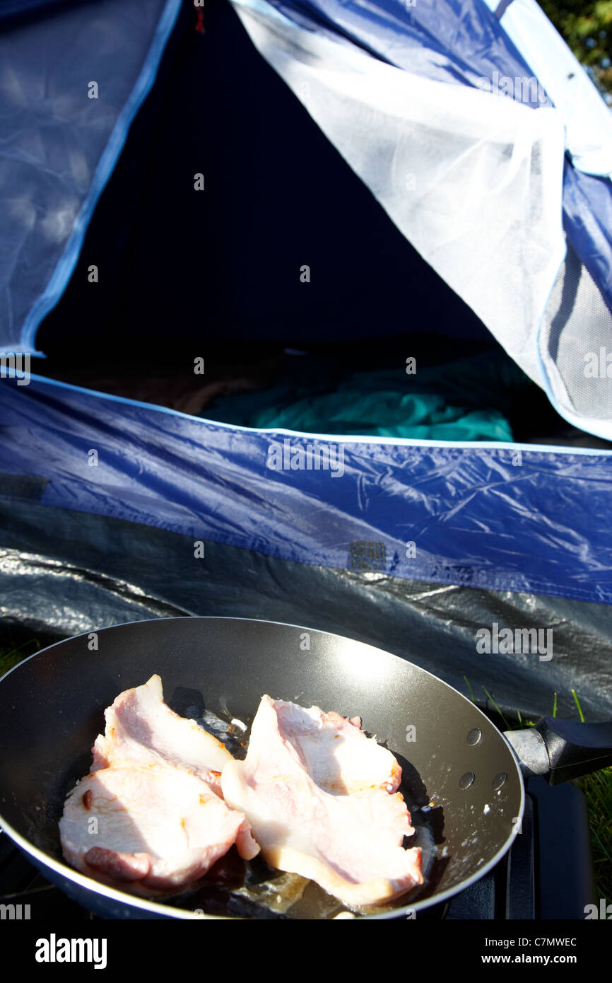 frying bacon on a small portable gas cooker in front of an open tent ...